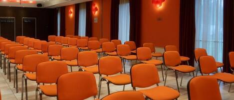 Uma sala de conferências com fileiras de cadeiras laranja, palco e uma grande janela com cortinas.