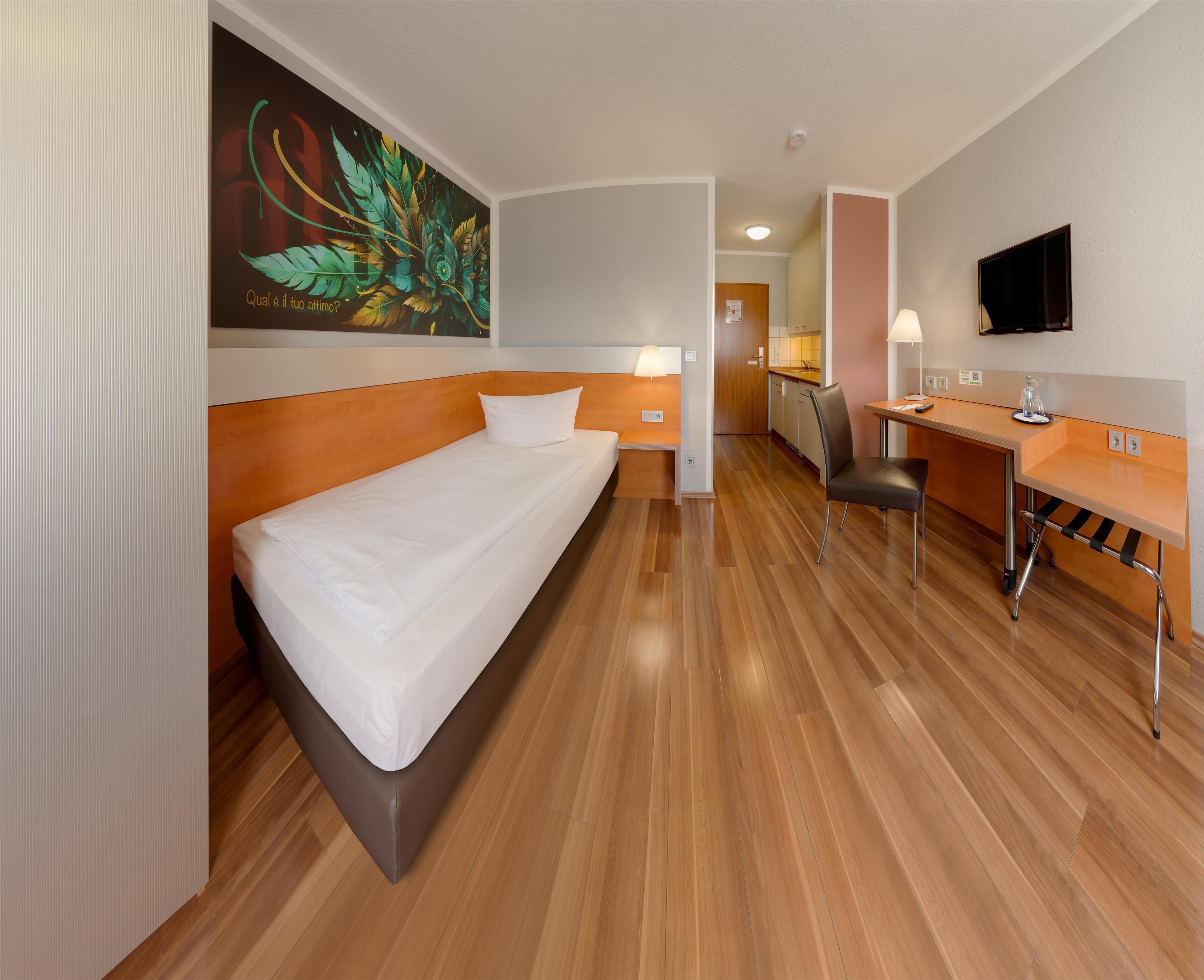 Quarto de hotel com uma cama de solteiro, uma escrivaninha, uma cadeira, uma televisão e um quadro na parede.