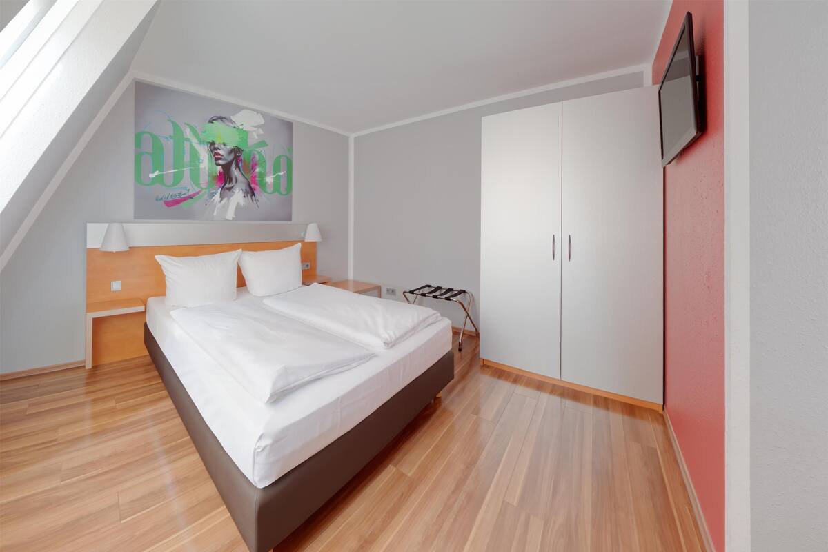 Quarto moderno com cama, mesa de cabeceira, guarda-roupa e televisão na parede.