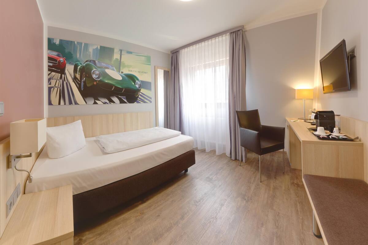 Quarto de hotel com cama, escrivaninha, cadeira, televisão e um quadro na parede.