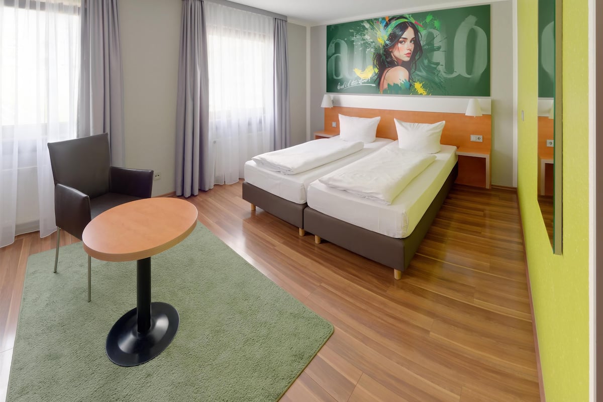 Quarto de hotel com duas camas, uma mesa redonda, uma cadeira e um quadro grande na parede.