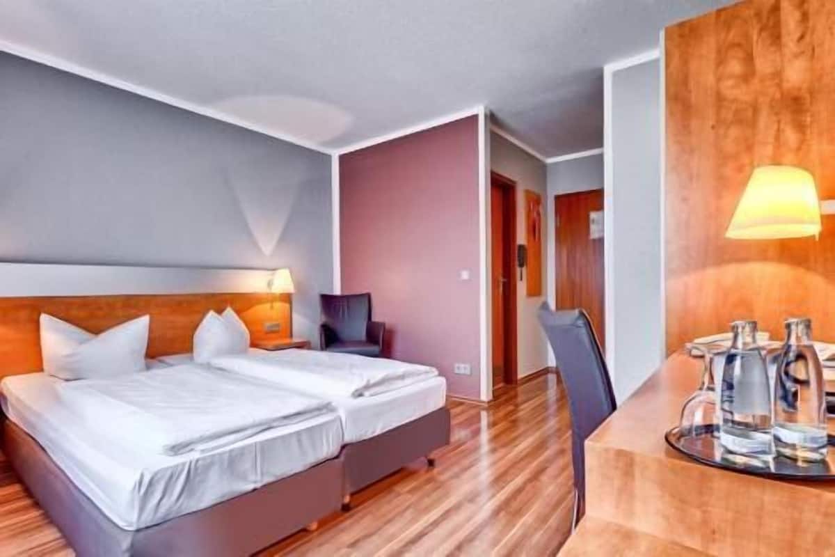 Quarto de hotel com uma cama de casal, uma escrivaninha com luminária e um aparador de madeira.
