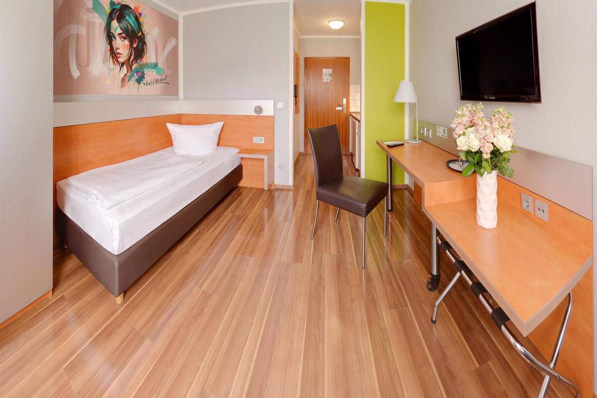 Quarto de hotel com cama, escrivaninha com cadeira, televisão e um quadro na parede.