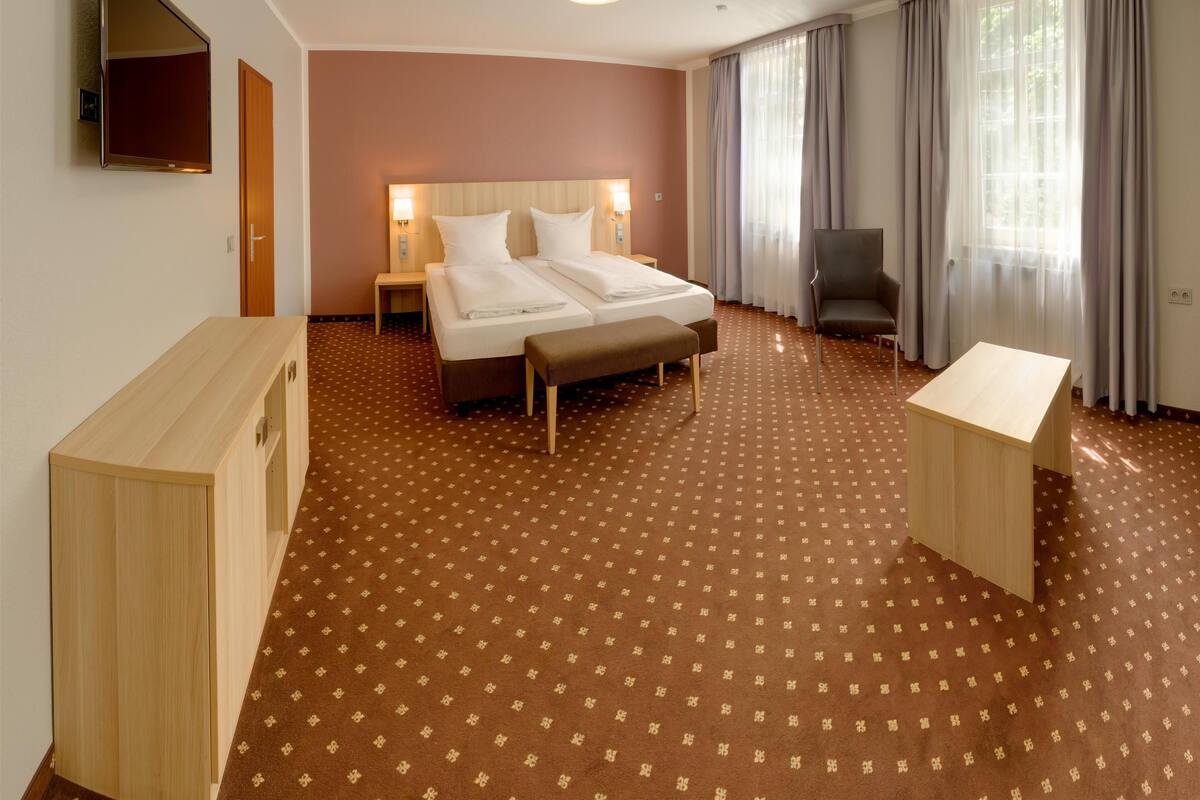 Quarto de hotel com cama, mesas de cabeceira, escrivaninha, cadeira e televisão fixada na parede.