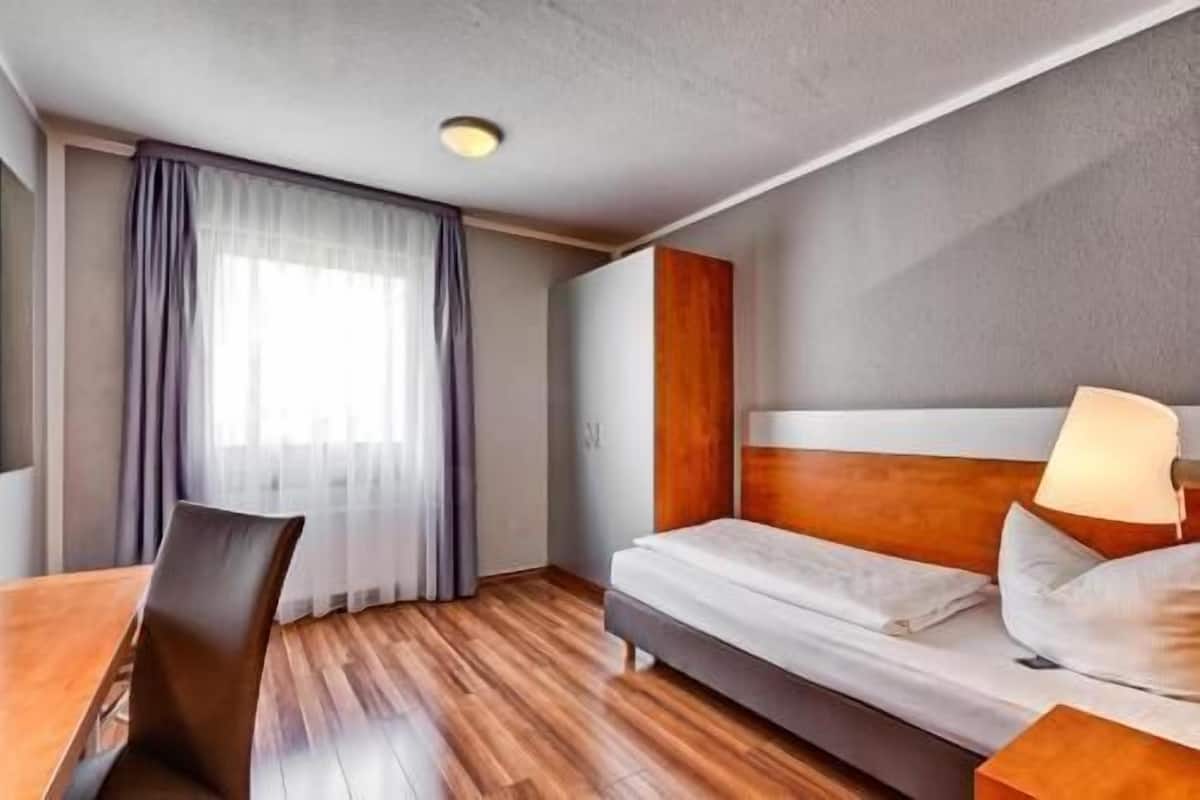 Quarto de hotel com piso de madeira, cama com roupa de cama branca, escrivaninha com cadeira e janela com cortinas leves.