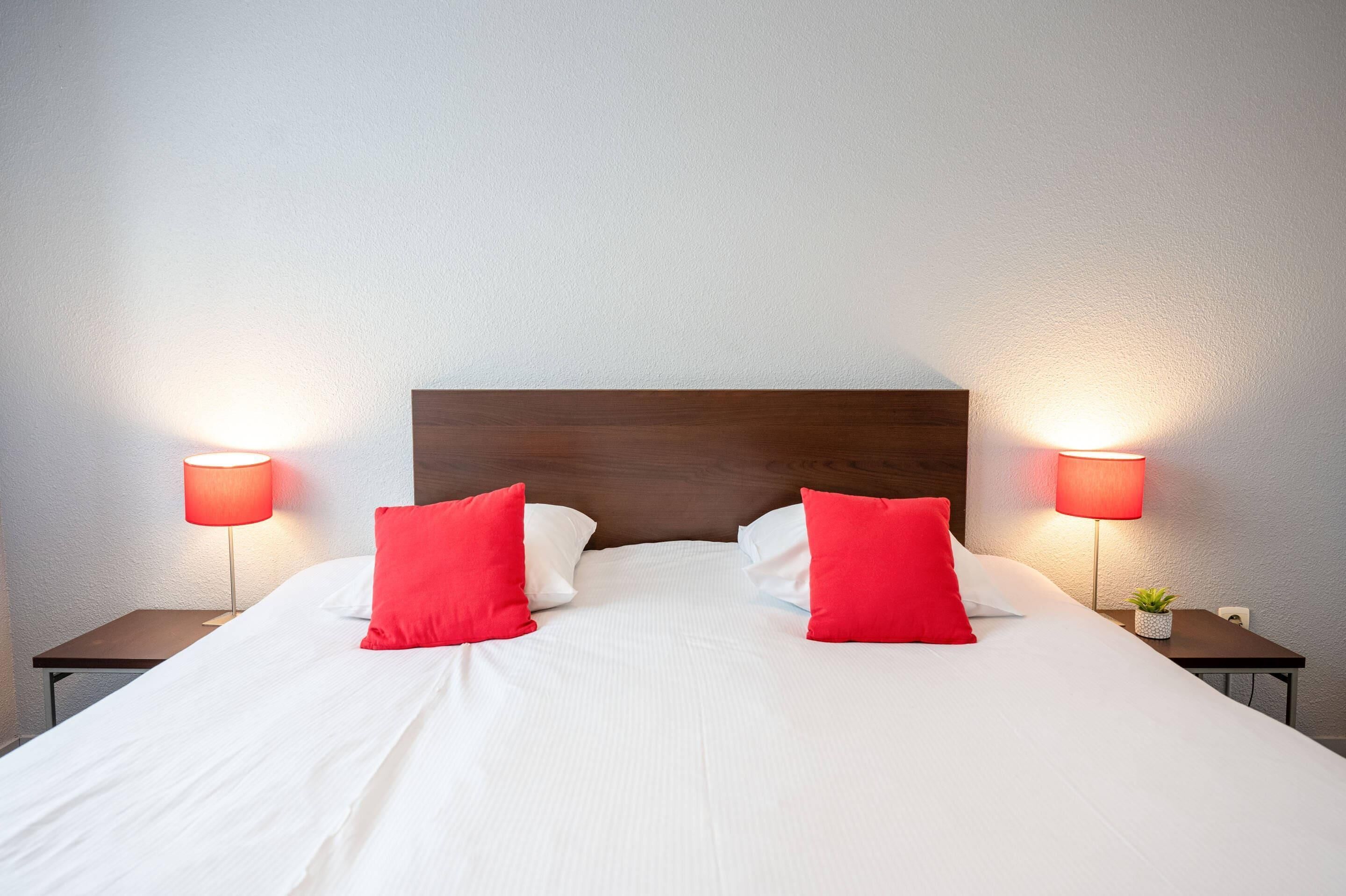 Cama bem arrumada com lençóis brancos e duas almofadas vermelhas, com duas mesas de cabeceira com abajures de cada lado, em frente a uma parede lisa.