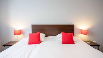 Cama bem arrumada com lençóis brancos e duas almofadas vermelhas, com duas mesas de cabeceira com abajures de cada lado, em frente a uma parede lisa.
