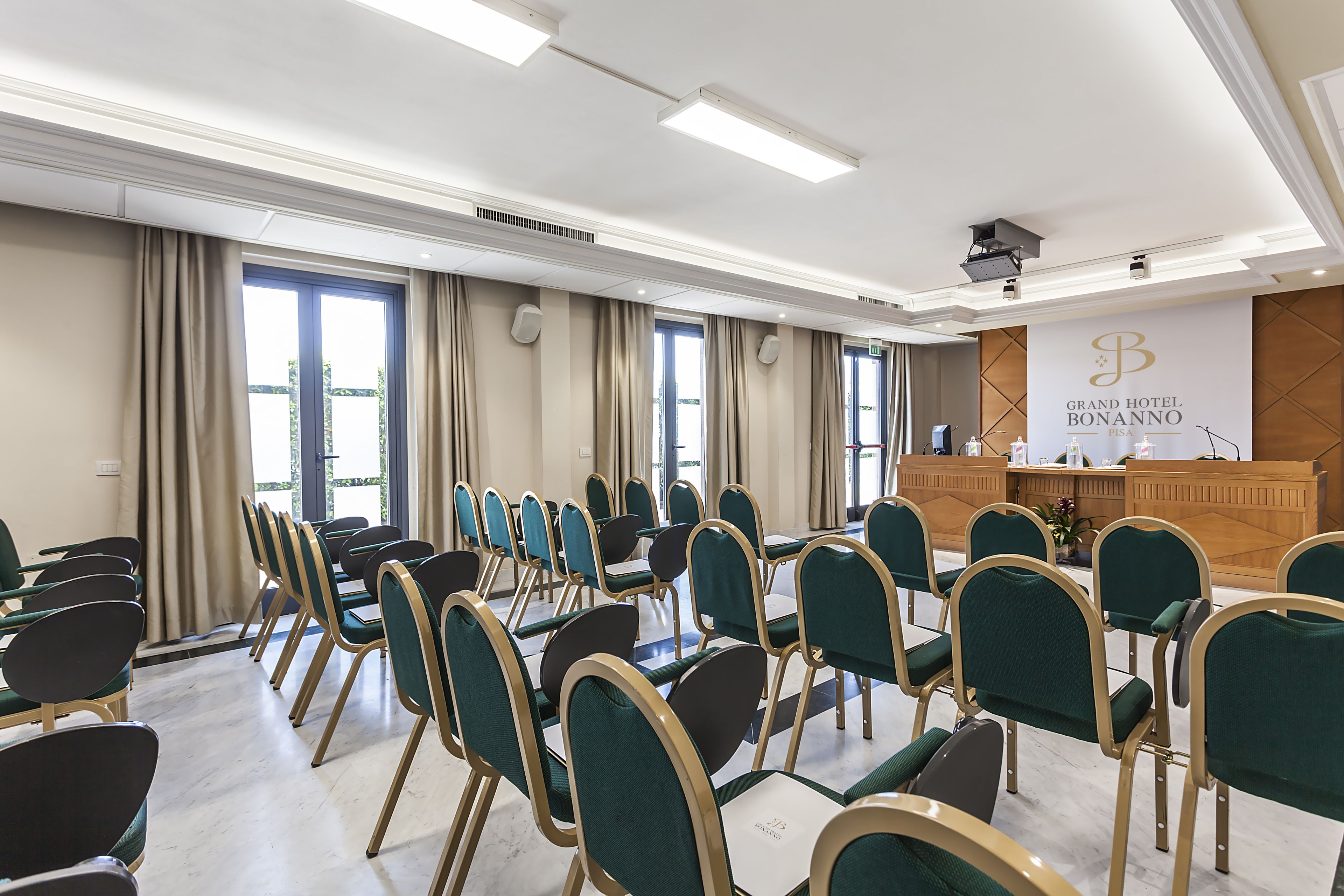 Uma sala de conferências com fileiras de cadeiras, um pódio e um banner com a inscrição 'Grand Hotel Bonanno'.