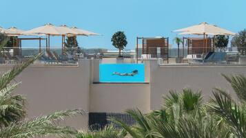 Uma área de piscina no terraço com espreguiçadeiras e guarda-sóis, céu azul e vista para o oceano.