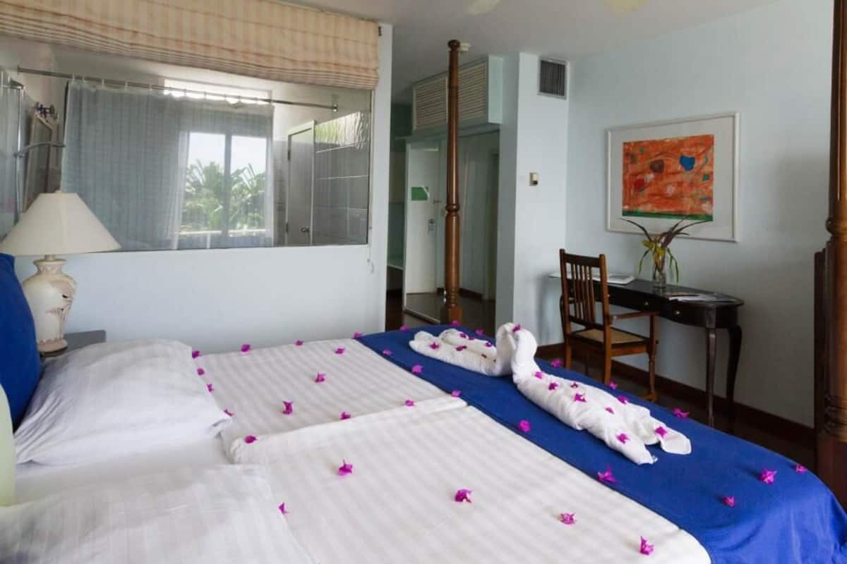 Quarto de hotel com duas camas, uma escrivaninha, uma cadeira, um abajur, janela com cortinas e um quadro na parede.