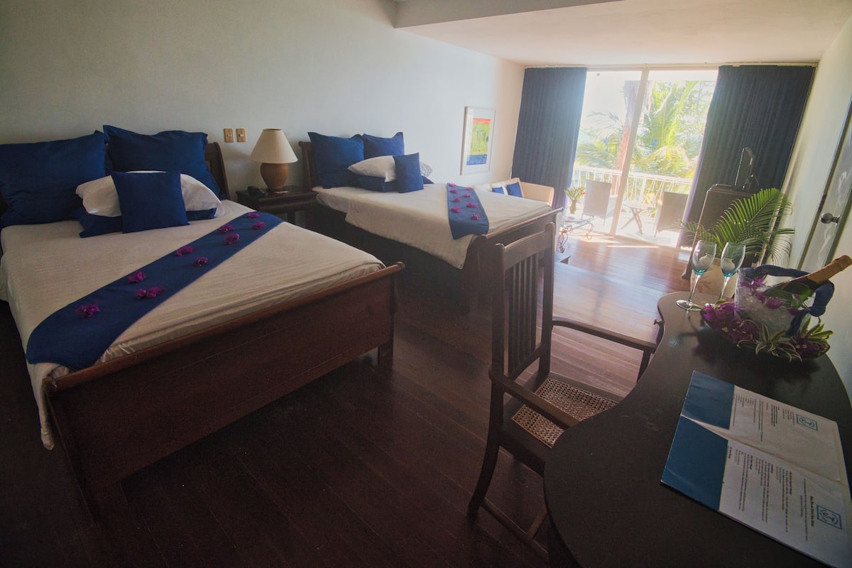 Quarto de hotel com duas camas, uma cadeira, uma mesa com garrafa e copos, e vista para o exterior.
