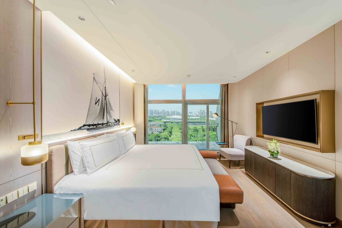Quarto de hotel moderno com uma cama grande, televisão, escrivaninha e vista para a cidade.