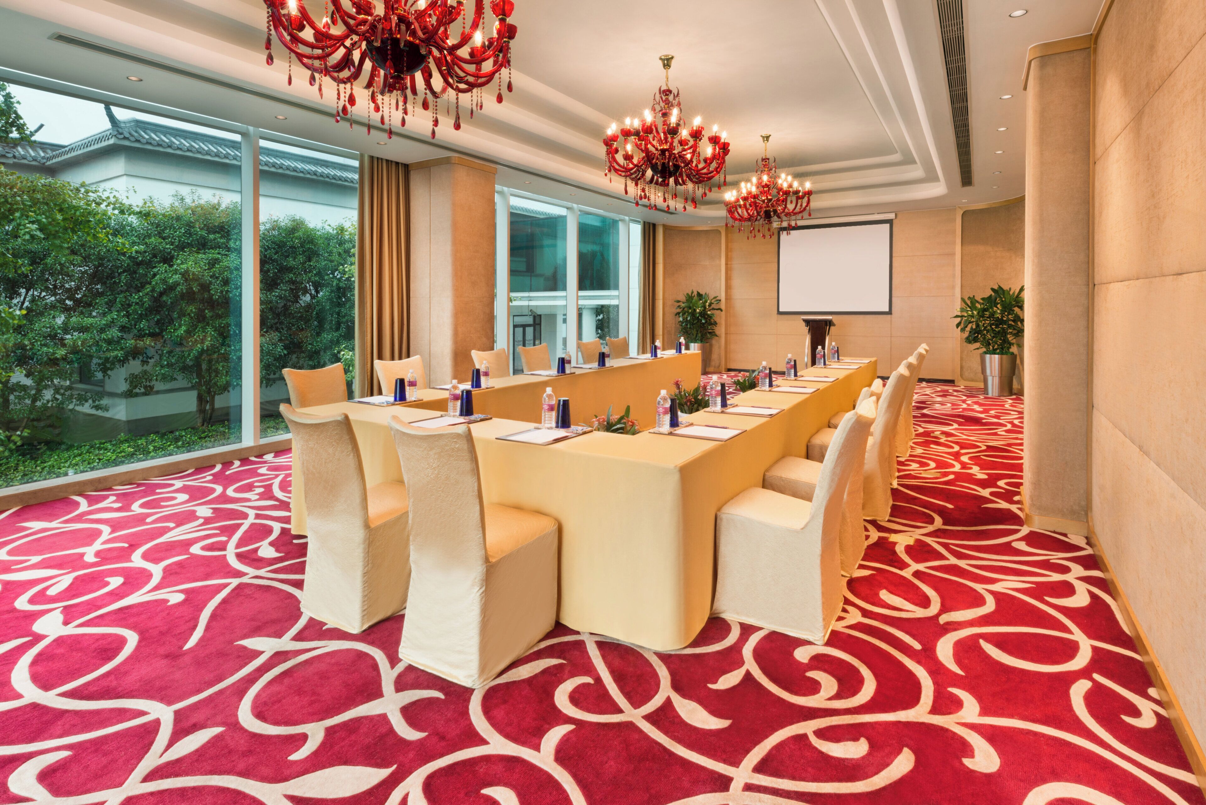 Sala de conferências com uma mesa grande, cadeiras, tela de projetor e tapete vermelho.