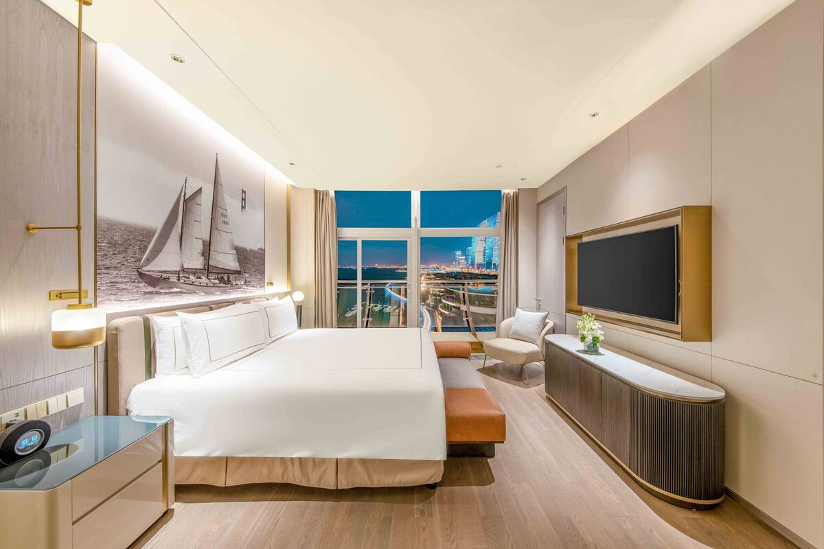 Quarto de hotel moderno com uma cama grande, televisão, escrivaninha e vista para o horizonte noturno.
