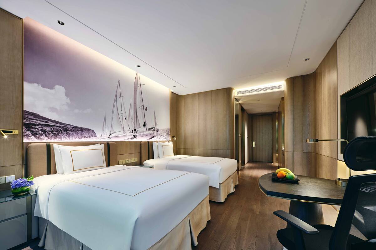 Quarto de hotel com duas camas, uma mesa, uma cadeira, uma TV e um mural com barcos à vela na parede.