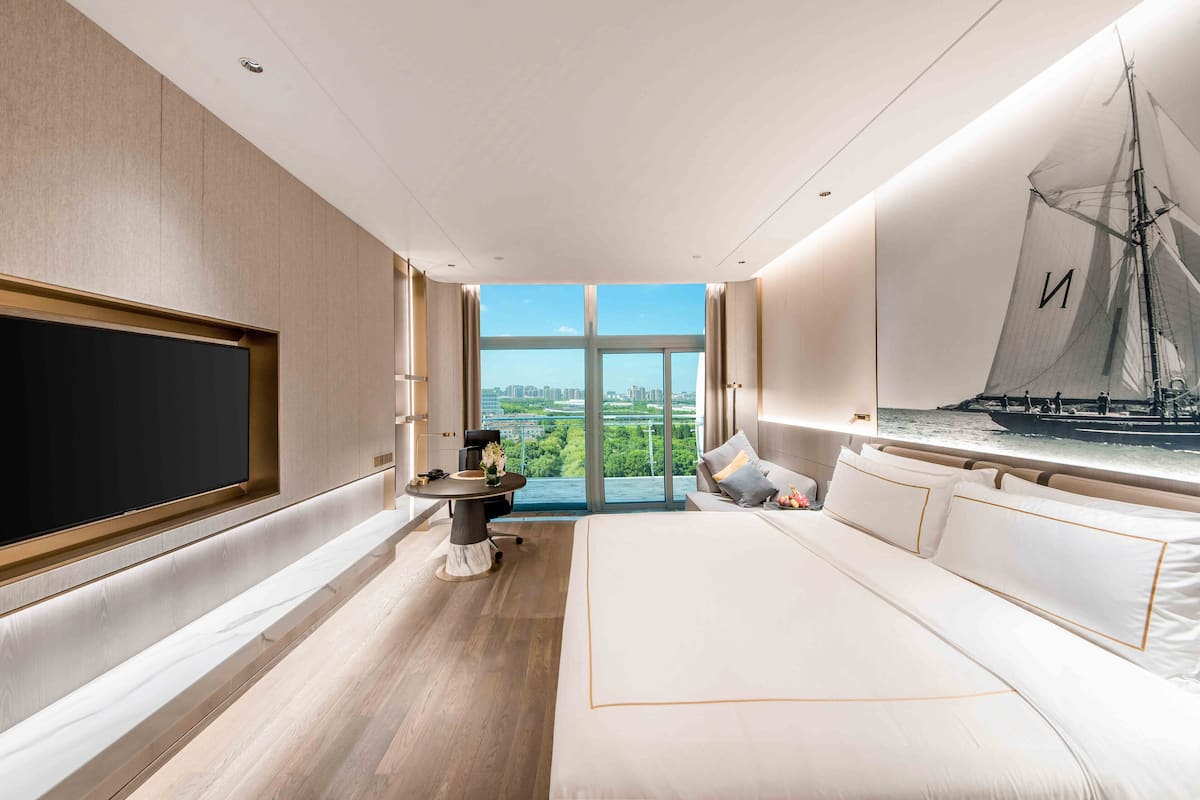 Quarto de hotel moderno com uma cama grande, televisão de tela plana, mesa de jantar e uma janela ampla com vista para a cidade.