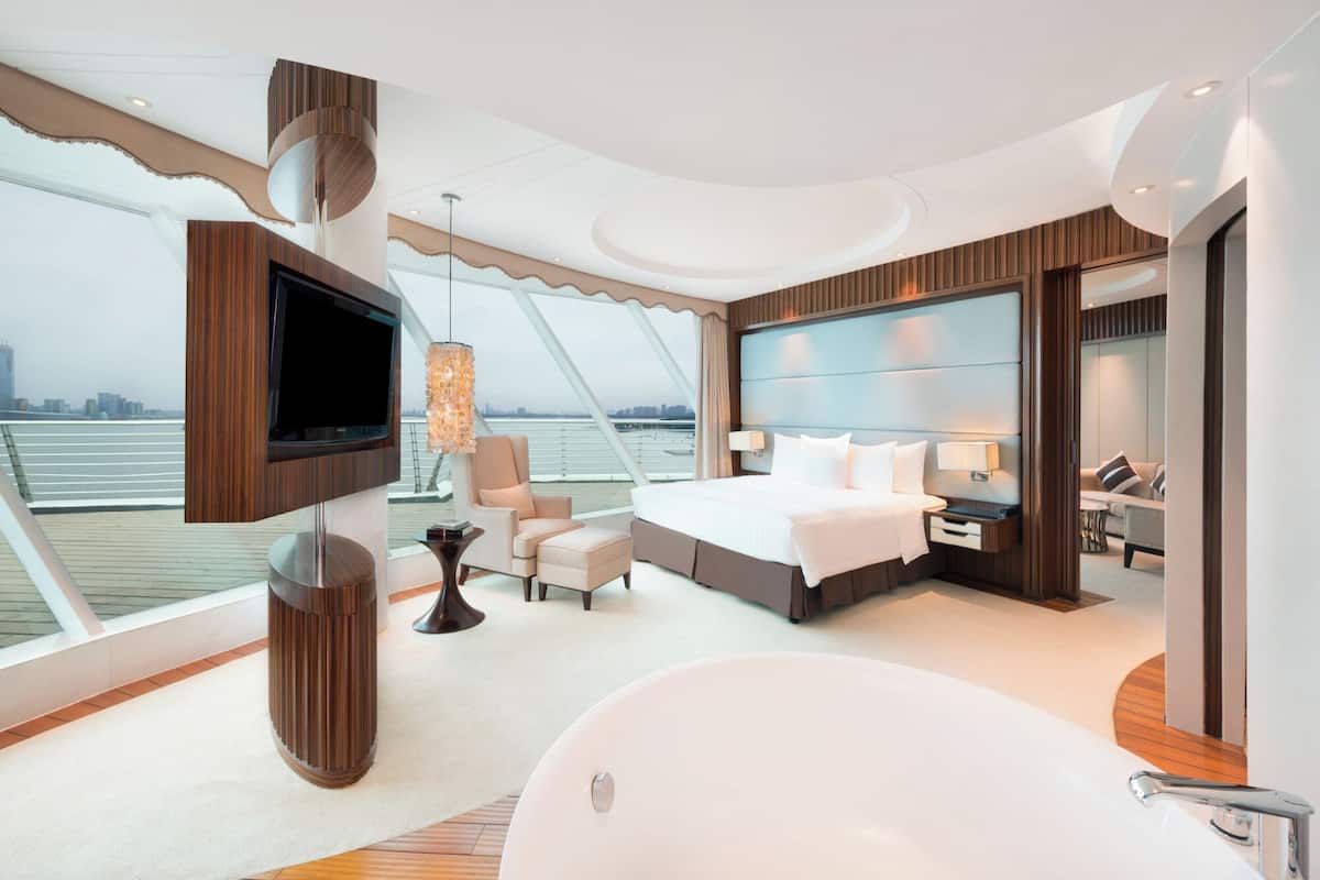 Quarto de hotel moderno com uma cama grande, uma televisão fixada na parede, área de estar com uma poltrona e uma mesa, e vista da cidade através de amplas janelas.