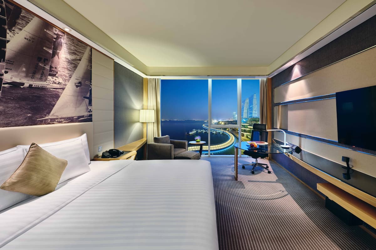 Quarto de hotel com uma cama grande, uma escrivaninha, uma cadeira, uma televisão e vista para a paisagem urbana.