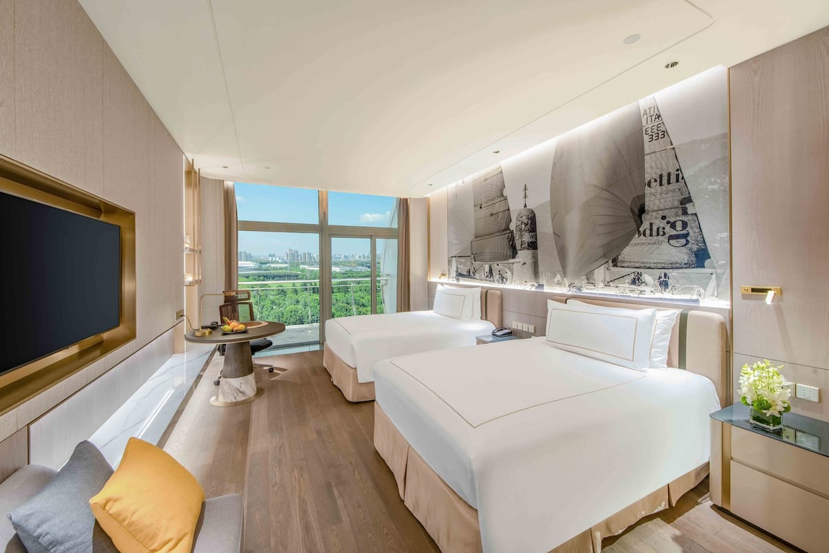Quarto de hotel moderno com duas camas, televisão, área de refeições e uma janela grande com vista para a cidade.
