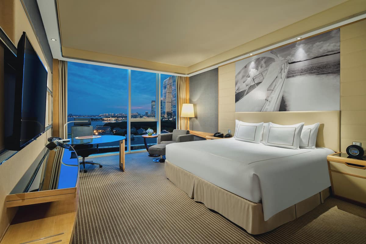 Quarto de hotel com uma cama grande, uma escrivaninha, uma cadeira, uma televisão e vista para a paisagem urbana.