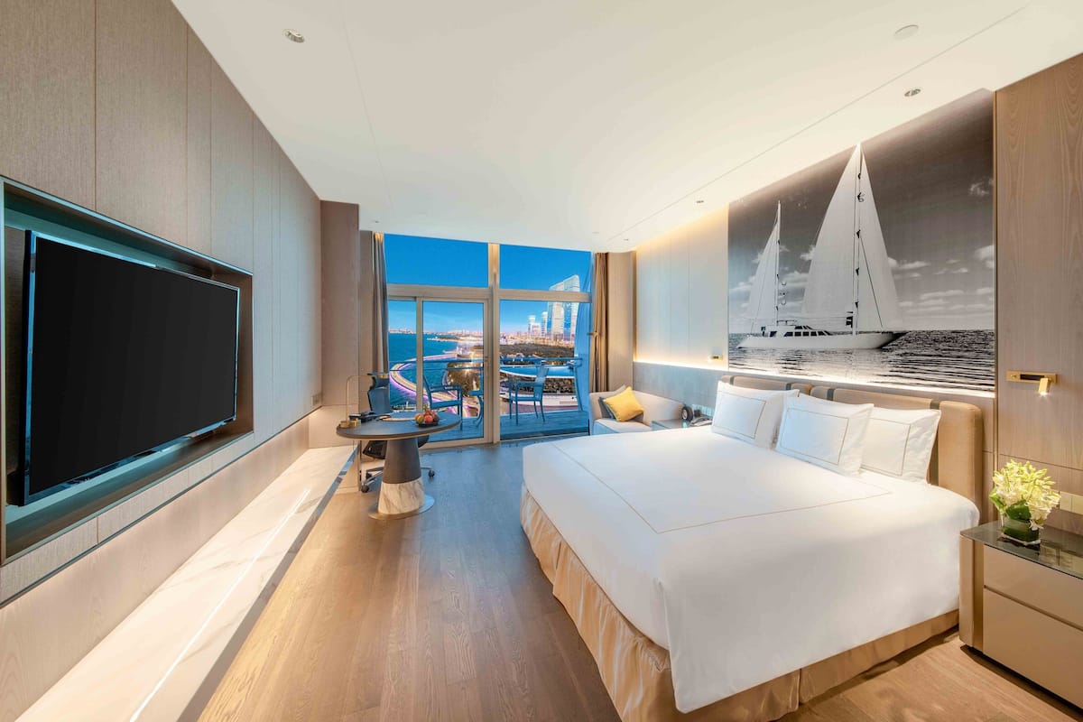 Quarto de hotel moderno com uma cama grande, uma televisão de tela plana, área de jantar com vista para a cidade e uma janela ampla.