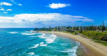 Uma praia costeira com ondas do oceano, areal e edifícios ao fundo.