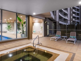 Uma área interna de piscina moderna com cadeiras de descanso, uma parede de vidro e vista para um edifício com vários andares.