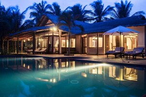Área junto à piscina com espreguiçadeiras, um espaço coberto para relaxar e um edifício com grandes janelas.