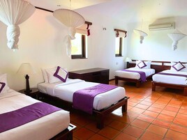 Quarto de hotel com quatro camas, cada uma com roupa de cama branca e detalhes em roxo, uma mesa de cabeceira de madeira, uma escrivaninha e uma janela com cortinas.