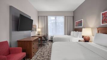Quarto de hotel com televisão de tela plana, uma escrivaninha com luminária, uma cadeira vermelha, duas camas e obras de arte nas paredes.