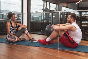 Duas pessoas sentadas em tapetes de exercício em uma academia, com equipamentos de ginástica ao fundo.