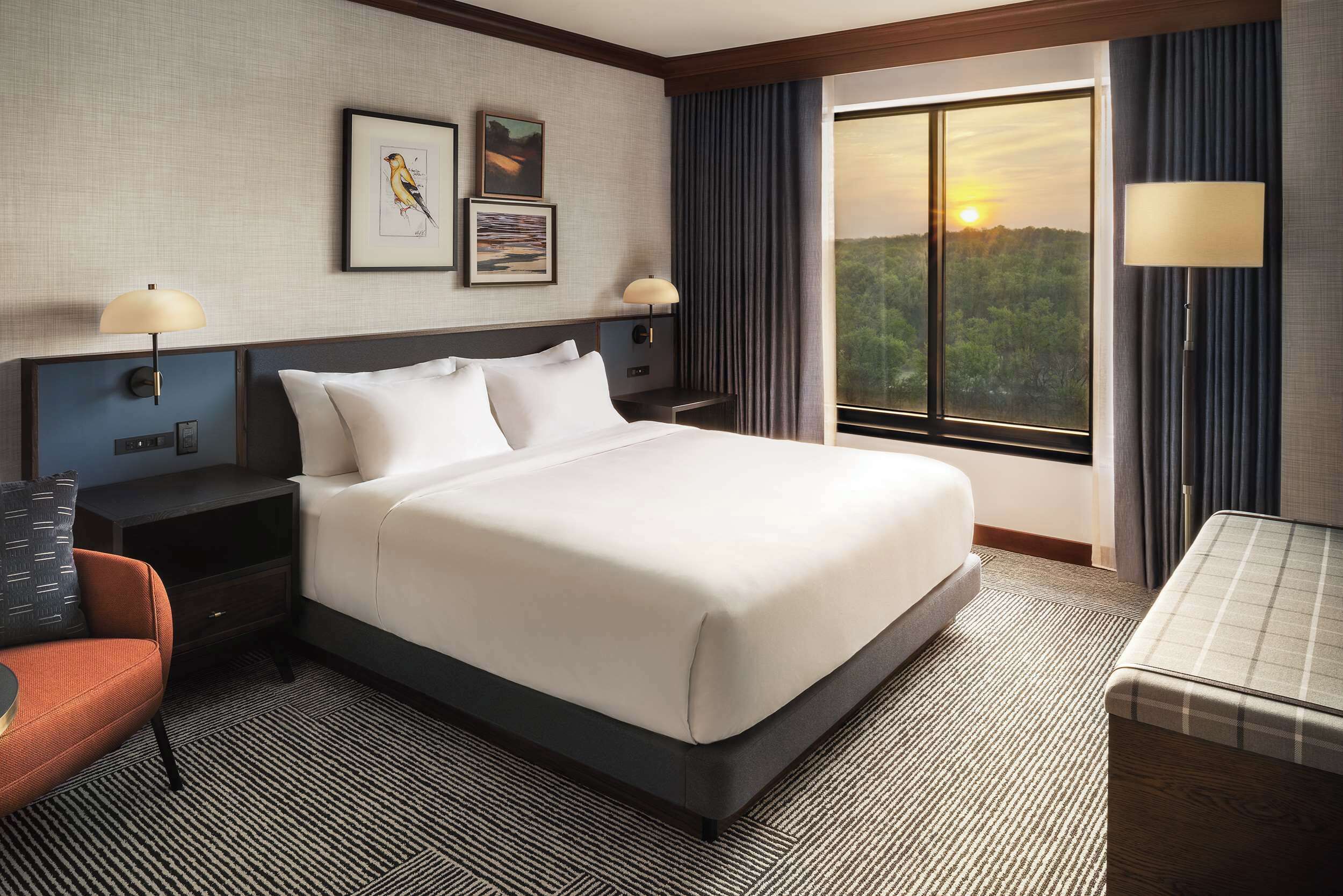 Quarto de hotel com uma cama grande, duas mesas de cabeceira, uma cadeira, uma escrivaninha e uma janela ampla com vista para o exterior.