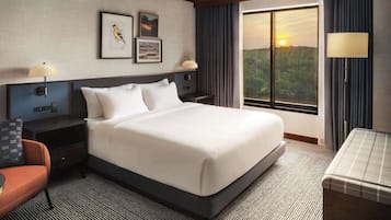 Quarto de hotel com uma cama grande, duas mesas de cabeceira, uma cadeira, uma escrivaninha e uma janela ampla com vista para o exterior.