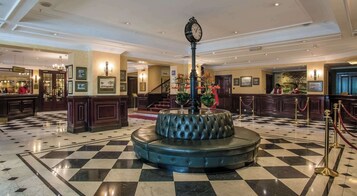 Um saguão com piso xadrez, área de estar circular central, um relógio e um balcão de recepção.