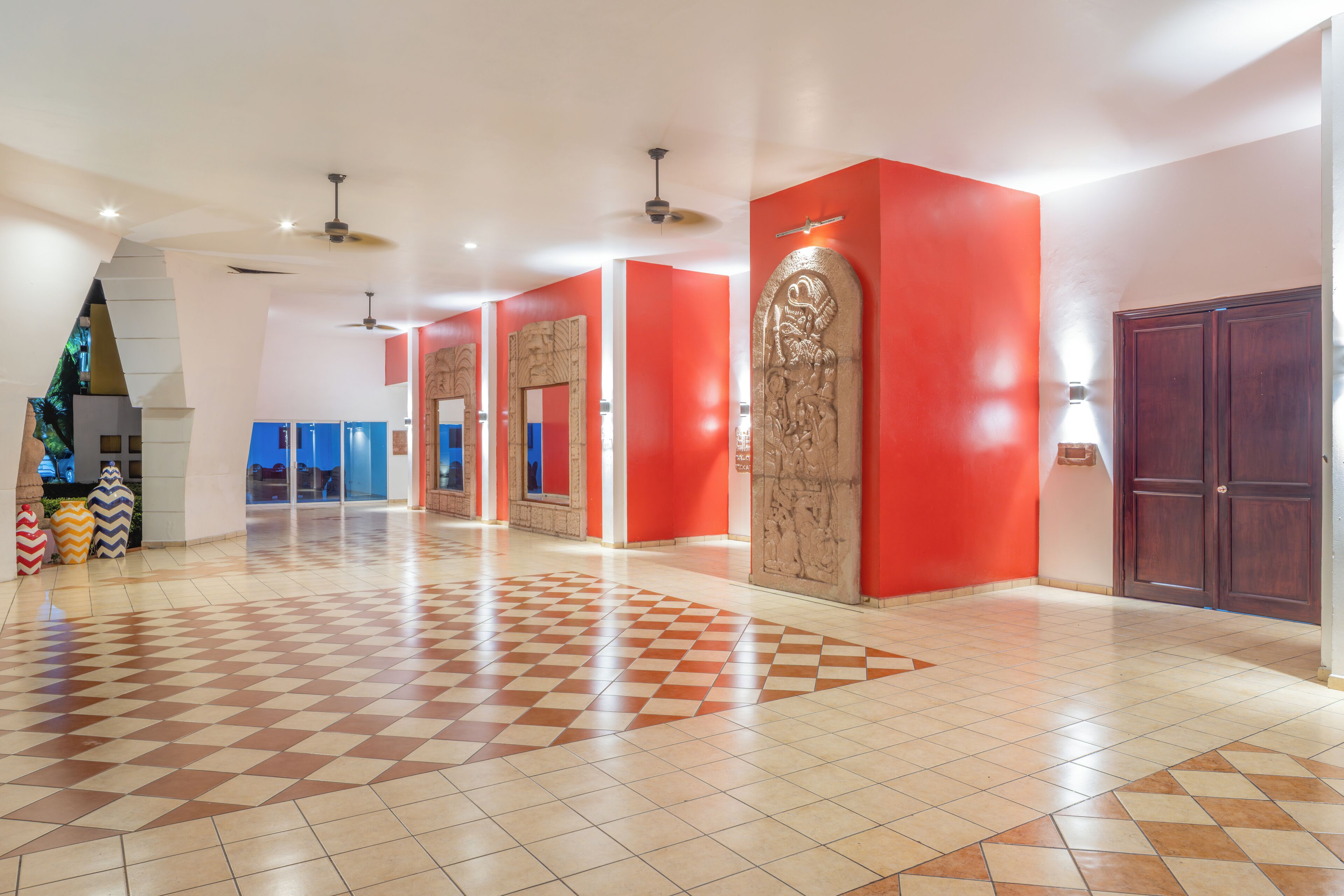 Um hall de hotel espaçoso com piso xadrez, paredes vermelhas e uma grande porta de madeira.