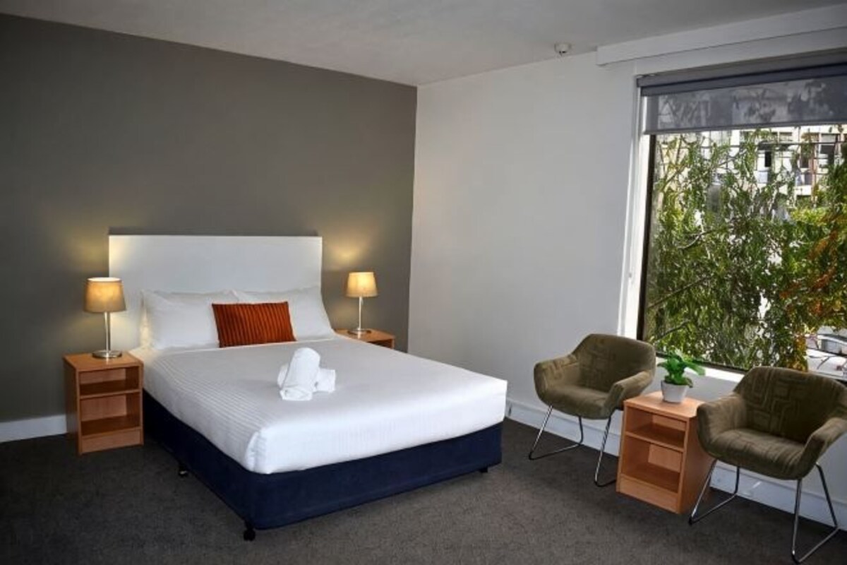 Quarto de hotel com uma cama grande, duas mesas de cabeceira, uma cadeira, uma pequena mesa com uma planta e uma janela com vista para árvores.