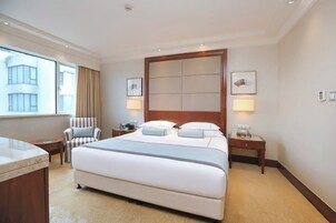Quarto de hotel com uma cama grande, uma cadeira, uma escrivaninha e uma janela com cortinas.