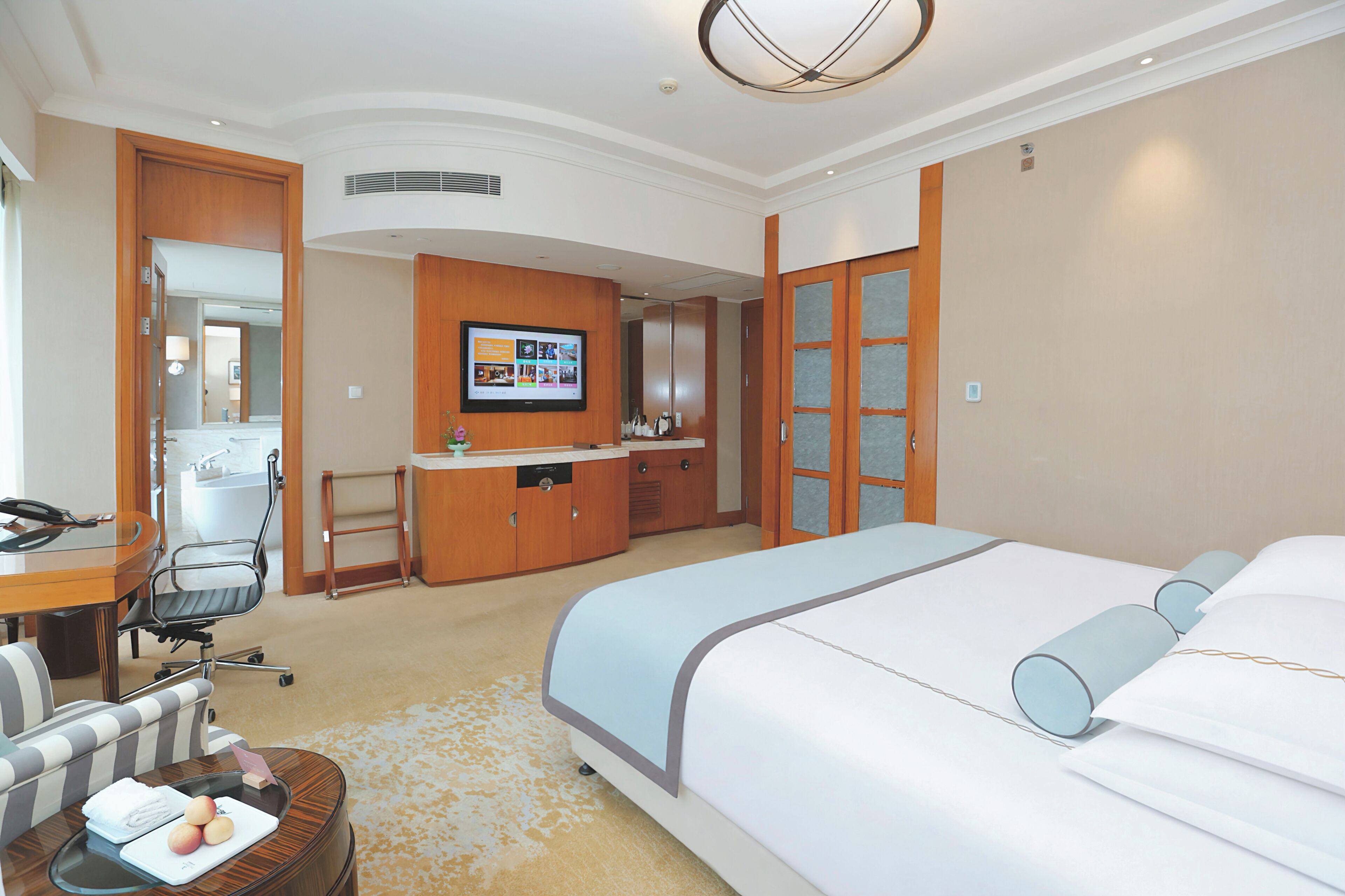 Quarto de hotel com uma cama grande, uma escrivaninha com cadeira, um espaço de estar pequeno e um banheiro visível por uma porta aberta.
