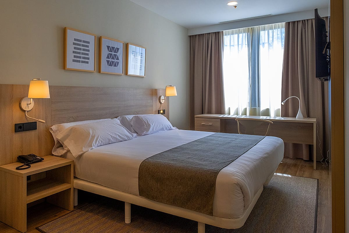 Quarto de hotel com cama, mesas de cabeceira, escrivaninha e televisão.