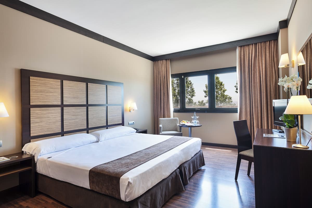Quarto de hotel com uma cama grande, uma escrivaninha com cadeira, uma mesinha e uma janela com cortinas.