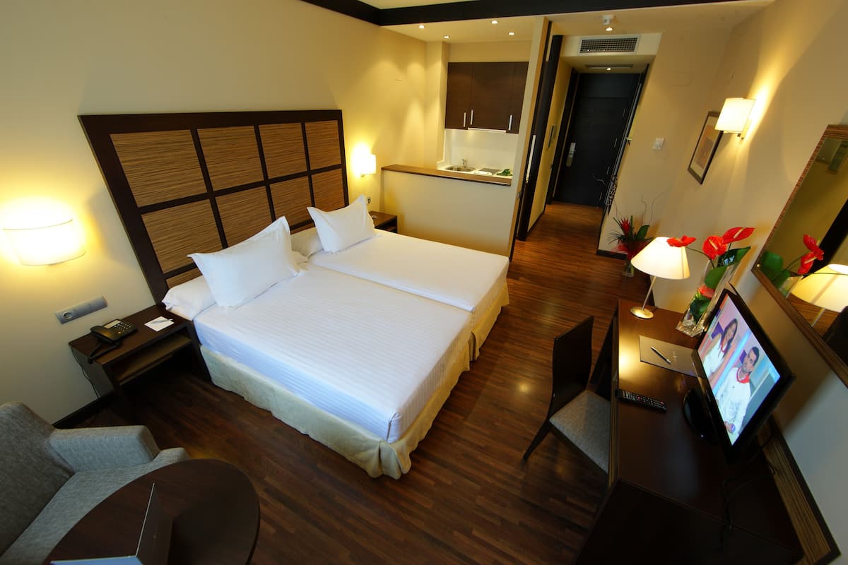 Quarto de hotel com uma cama grande, uma escrivaninha com televisão, uma cadeira, uma mesa de cabeceira e uma pequena área de cozinha.