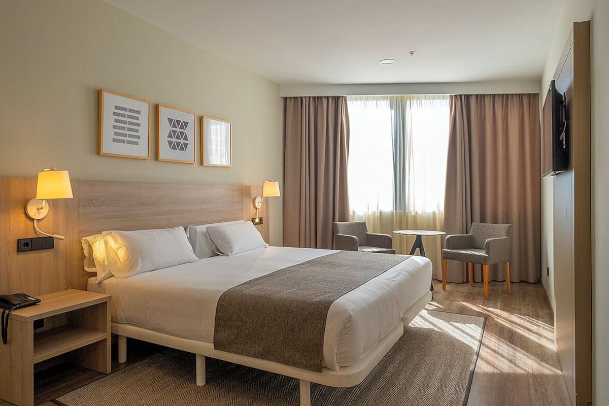 Quarto de hotel com uma cama grande, abajures, escrivaninha, cadeira e televisão na parede.