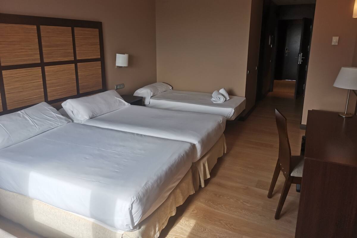 Quarto de hotel com duas camas, uma mesa, uma cadeira e uma porta que dá para outro espaço.