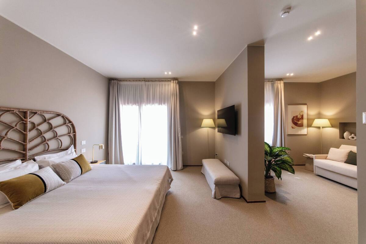 Quarto de hotel moderno com uma cama grande, área de estar com sofá e um banco, televisão de tela plana na parede e um vaso de planta.