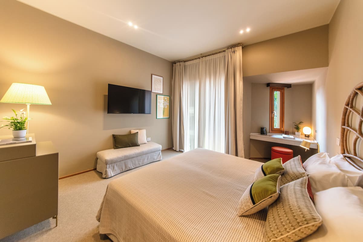 Quarto com uma cama grande, mesas de cabeceira, televisão, escrivaninha com cadeira e janela com cortinas.