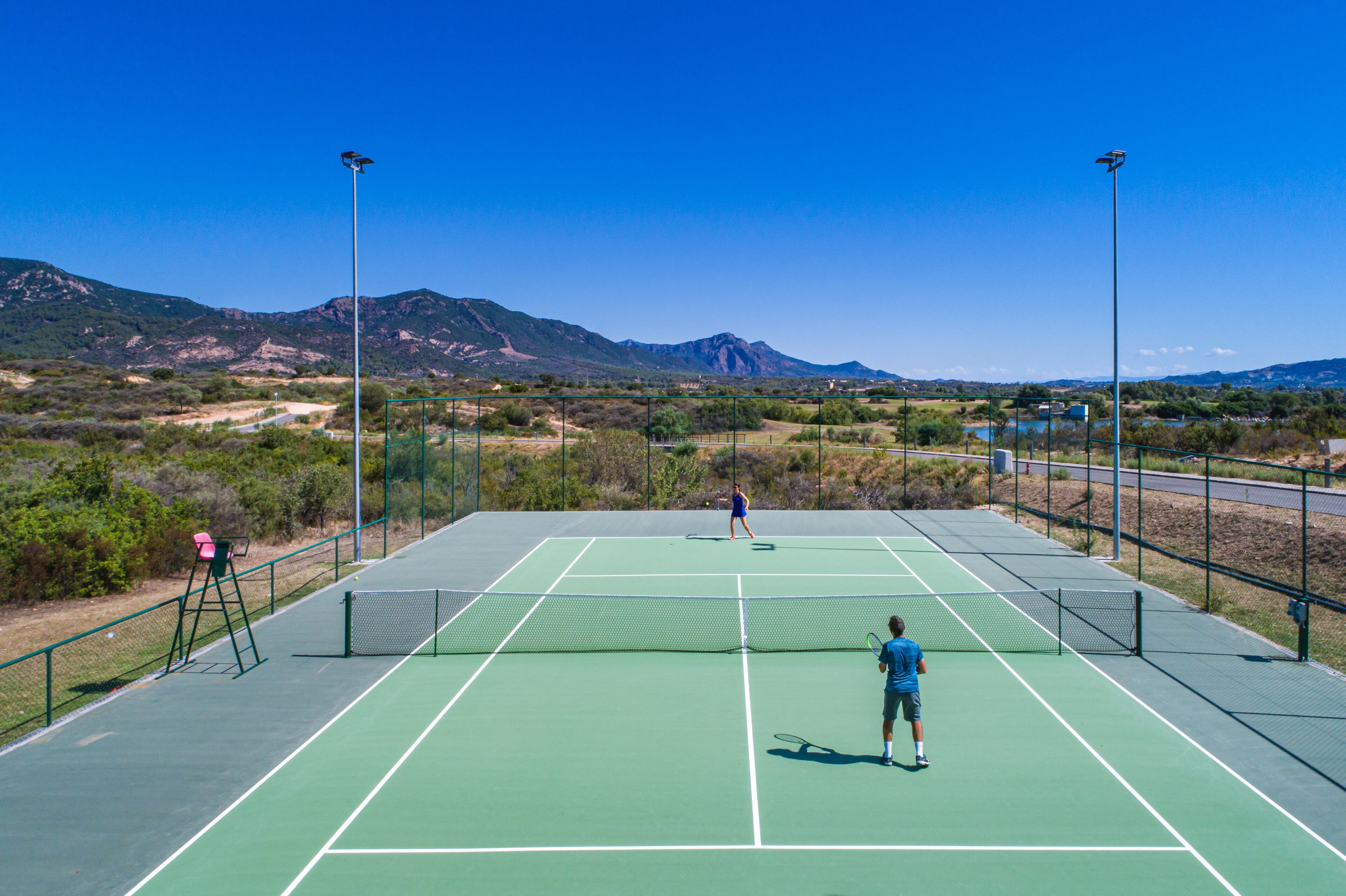 Campo de tênis ao ar livre com dois jogadores, cercado por montanhas e vegetação.