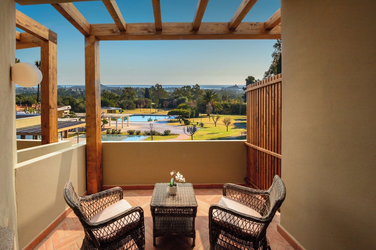 Varanda com cadeiras de vime e uma mesa, com vista para uma piscina e jardim ajardinado.