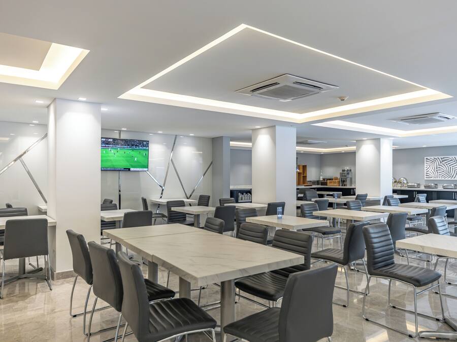 Uma sala de jantar moderna com várias mesas e cadeiras, uma tela grande exibindo um evento esportivo e uma área de cozinha ao fundo.