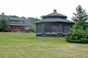 Um gazebo com telhado verde e estrutura de madeira, cercado por um gramado e um edifício vermelho ao fundo.