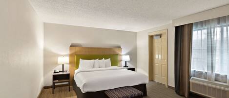 Quarto de hotel com uma cama grande, duas mesas de cabeceira com luminárias, um banquinho no rodapé da cama, uma porta e uma janela com cortinas.